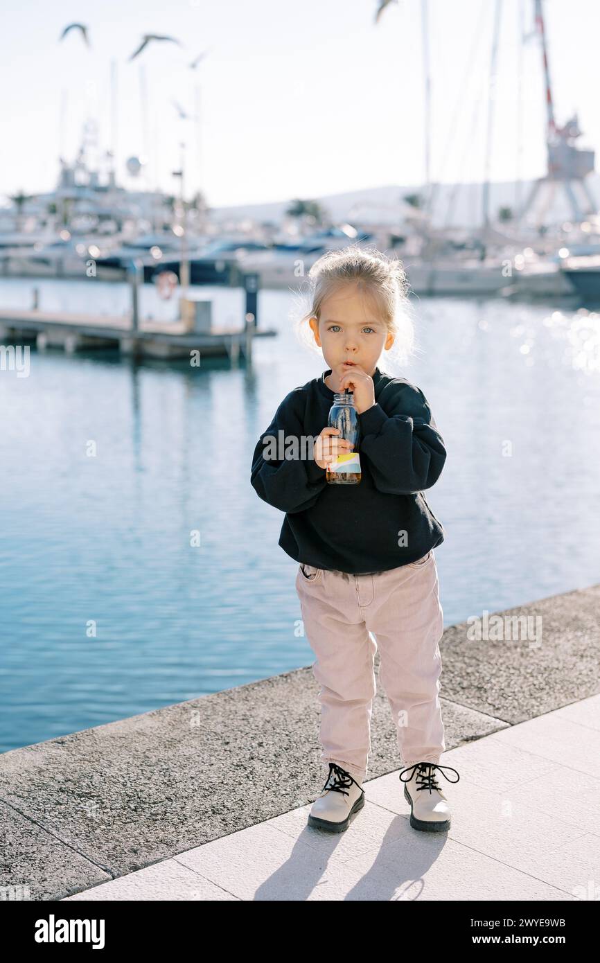 Little girl drinks juice through a straw from a bottle while standing ...