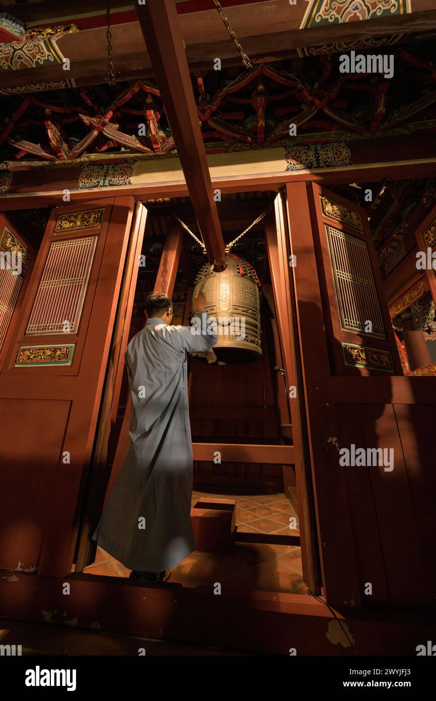 A monk in traditional attire ringing a large bell in Baoan temple ...