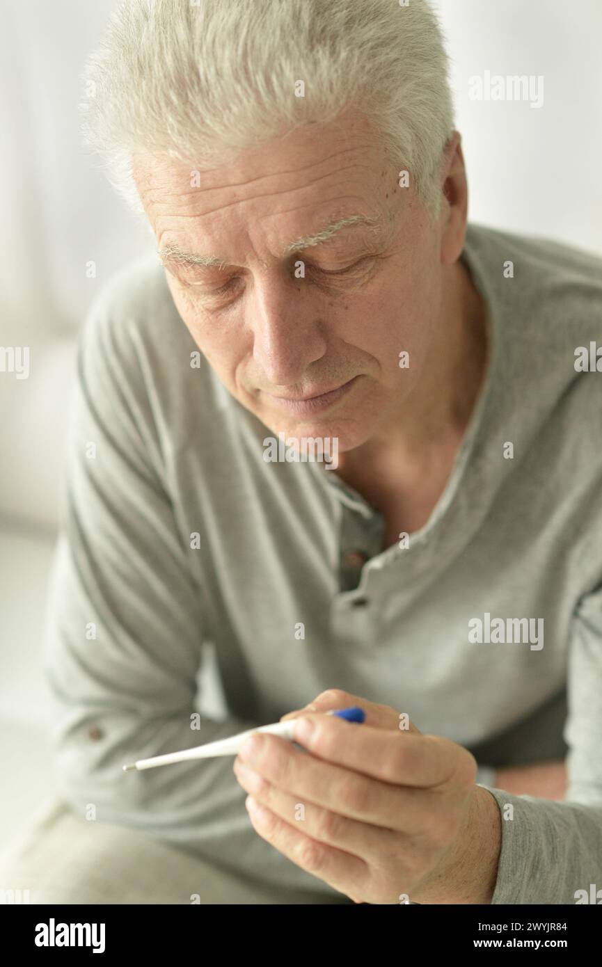 A sick elderly man sits on his bed and checks his body temperature ...