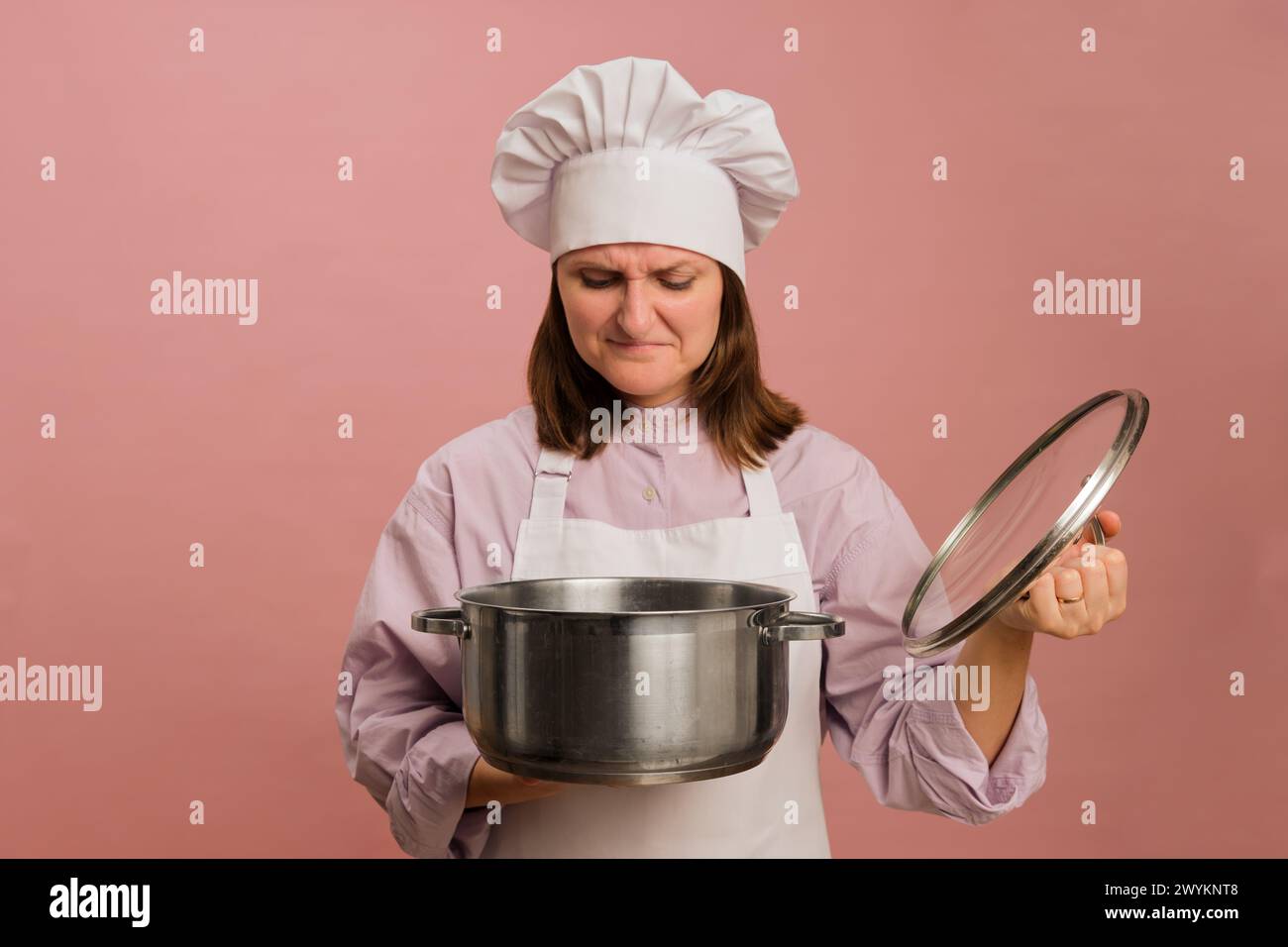 Woman cook looking with disgust into a pan on a studio pink background ...