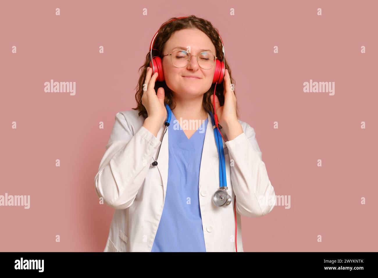 Woman doctor listening to music on headphones, studio pink background ...