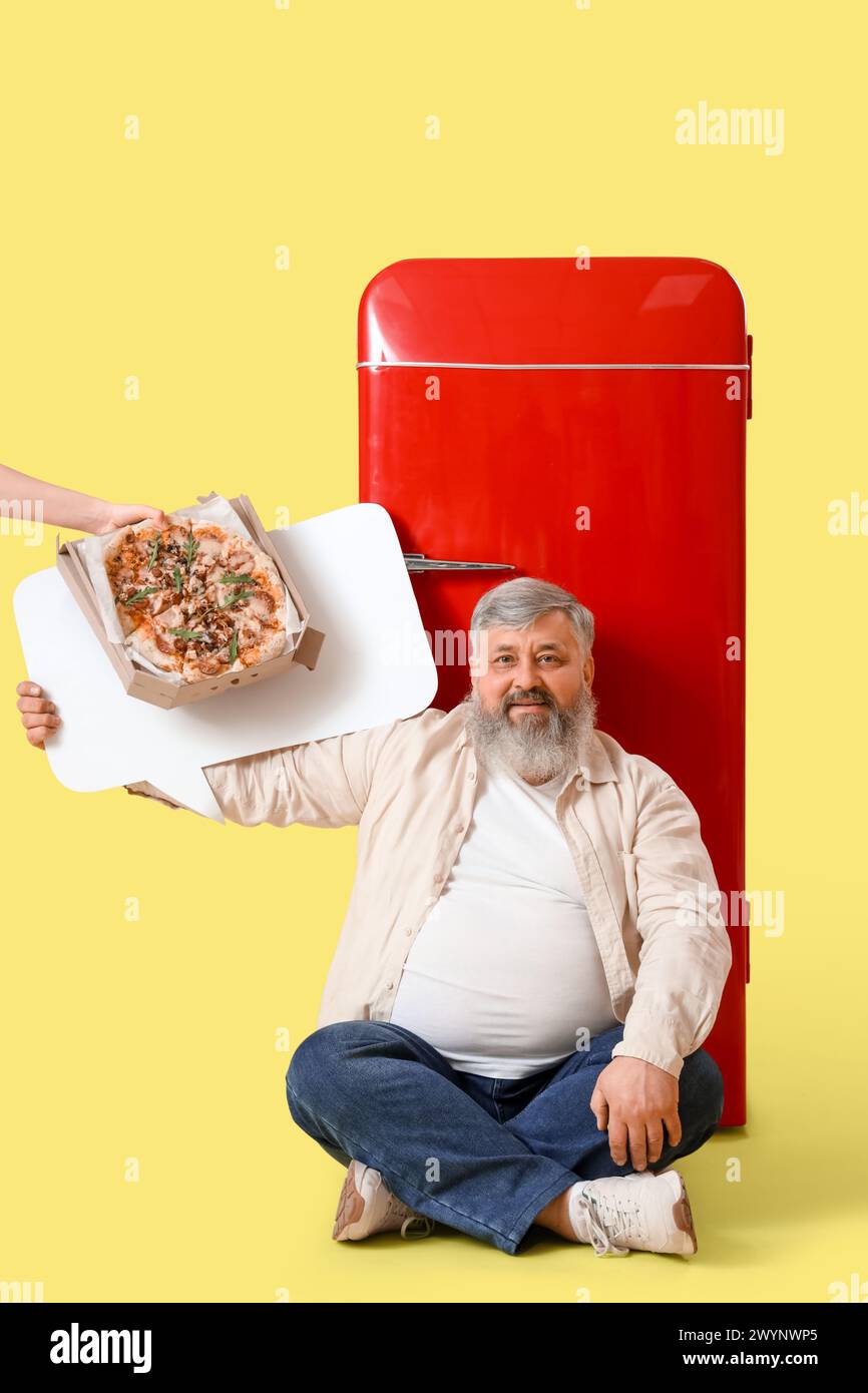 Overweight mature man with pizza sitting near fridge on yellow ...
