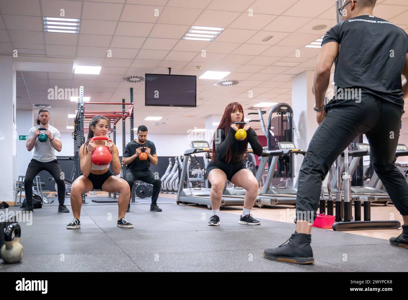 A diverse group of gym-goers engages in a kettlebell squat session with ...