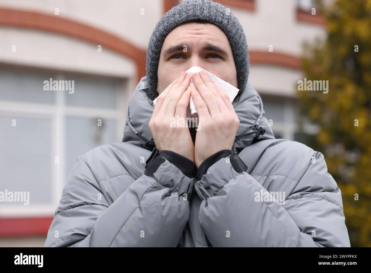 Sick young man with tissue blowing runny nose outdoors. Cold symptom ...