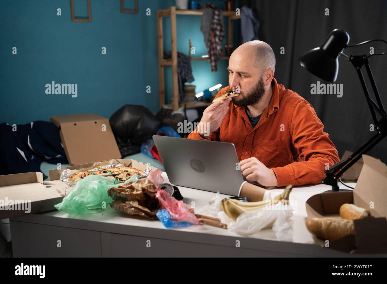 Bearded man eating pizza and working in messy, cluttered freelancer ...