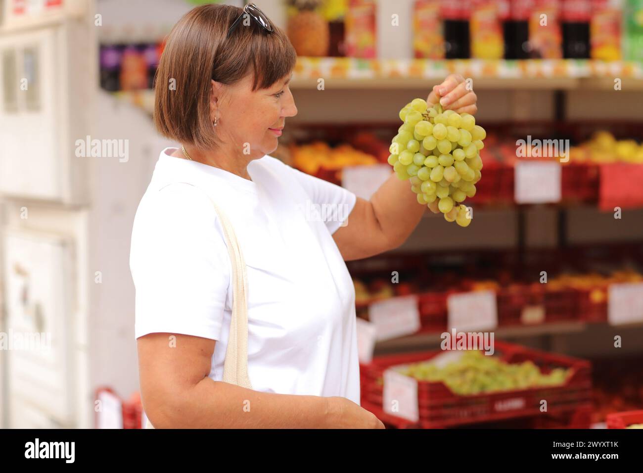 Middle aged woman holds branch of fresh yellow grapes, chooses and buys ...