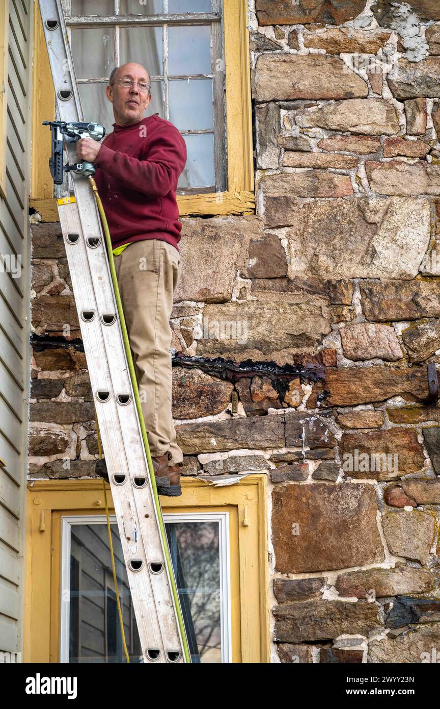 Man with pneumatic brad gun on a ladder for old stone home roof repair ...