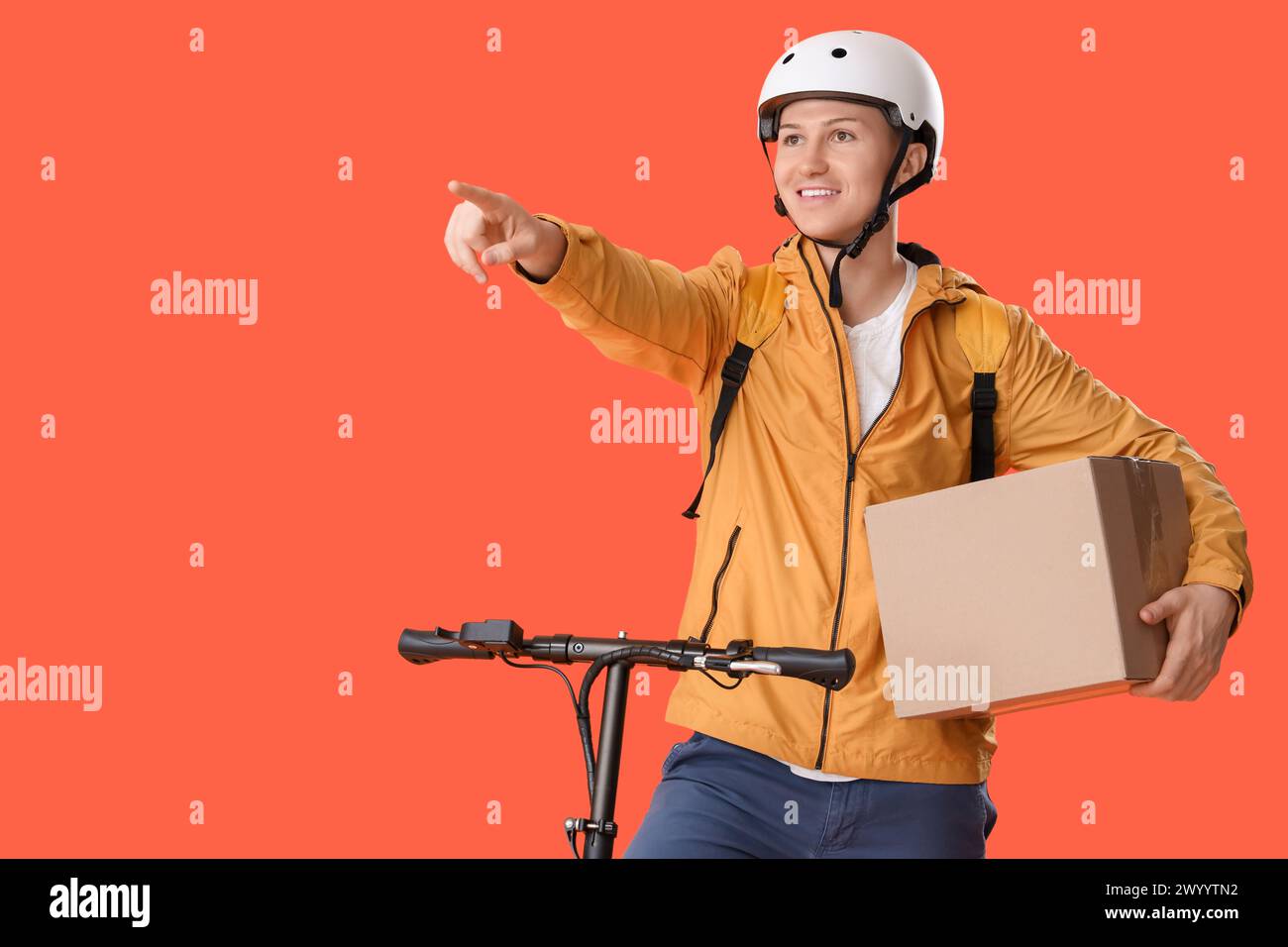 Male courier with cardboard box and kick scooter pointing at something ...