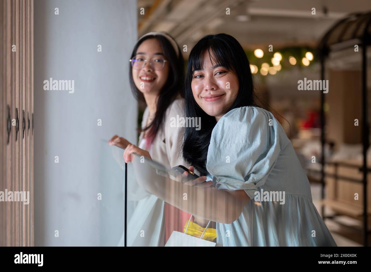 Two smiling young Asian women lean against a window, with shopping bags ...