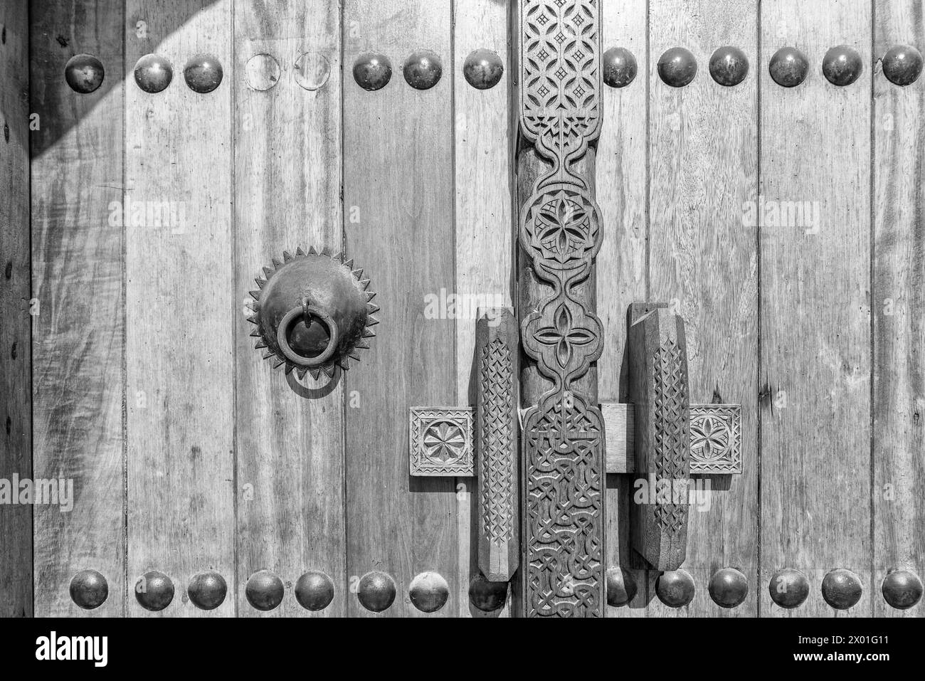 A monochrome image showing detail of a carved wooden door, handle and ...