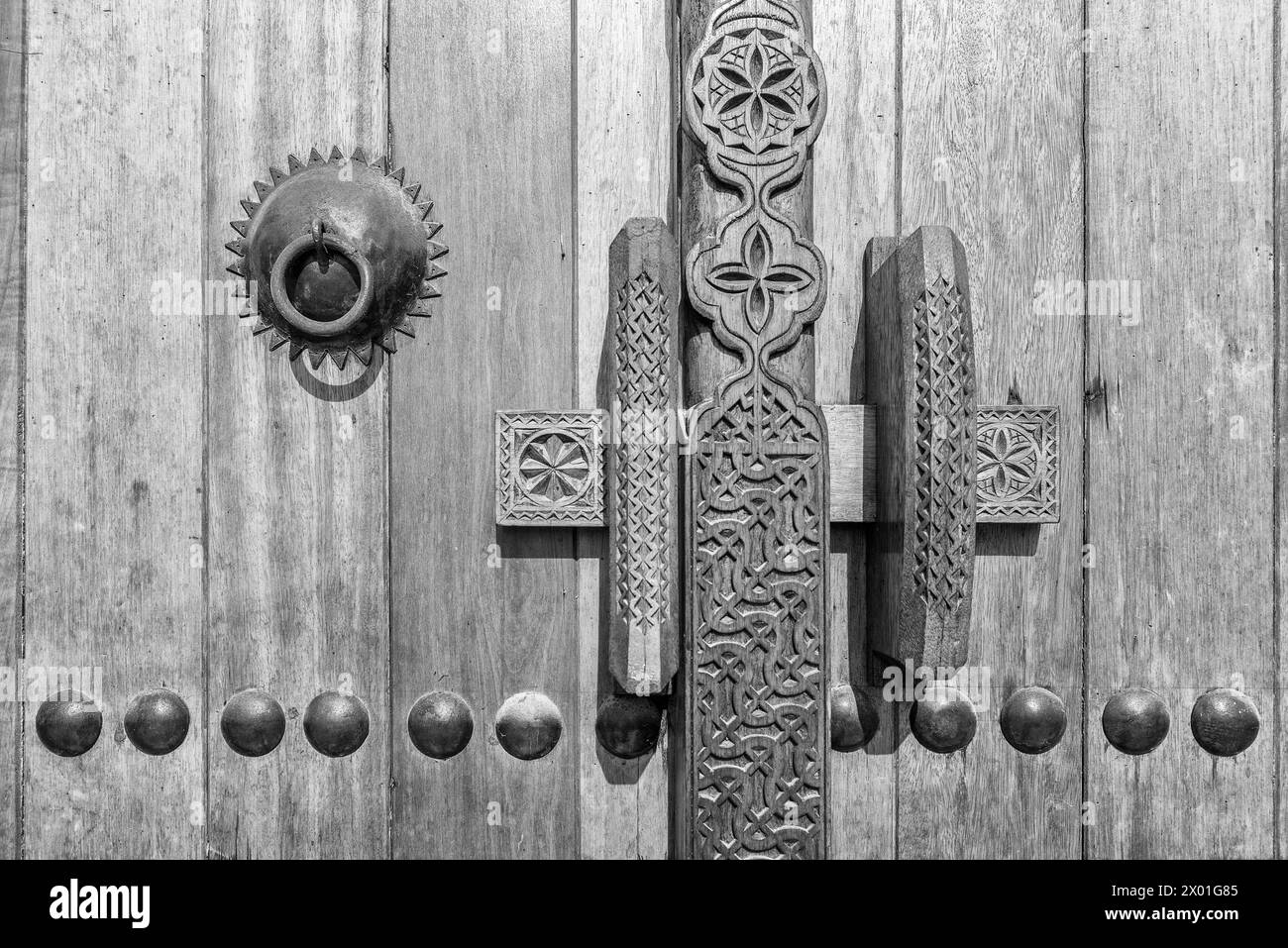 A monochrome image showing detail of a carved wooden door, handle and ...