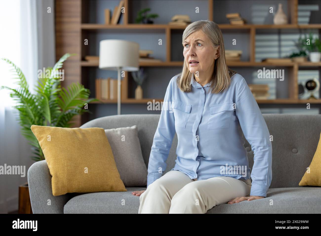Stressed female in casual outfit sitting on grey sofa and looking aside ...