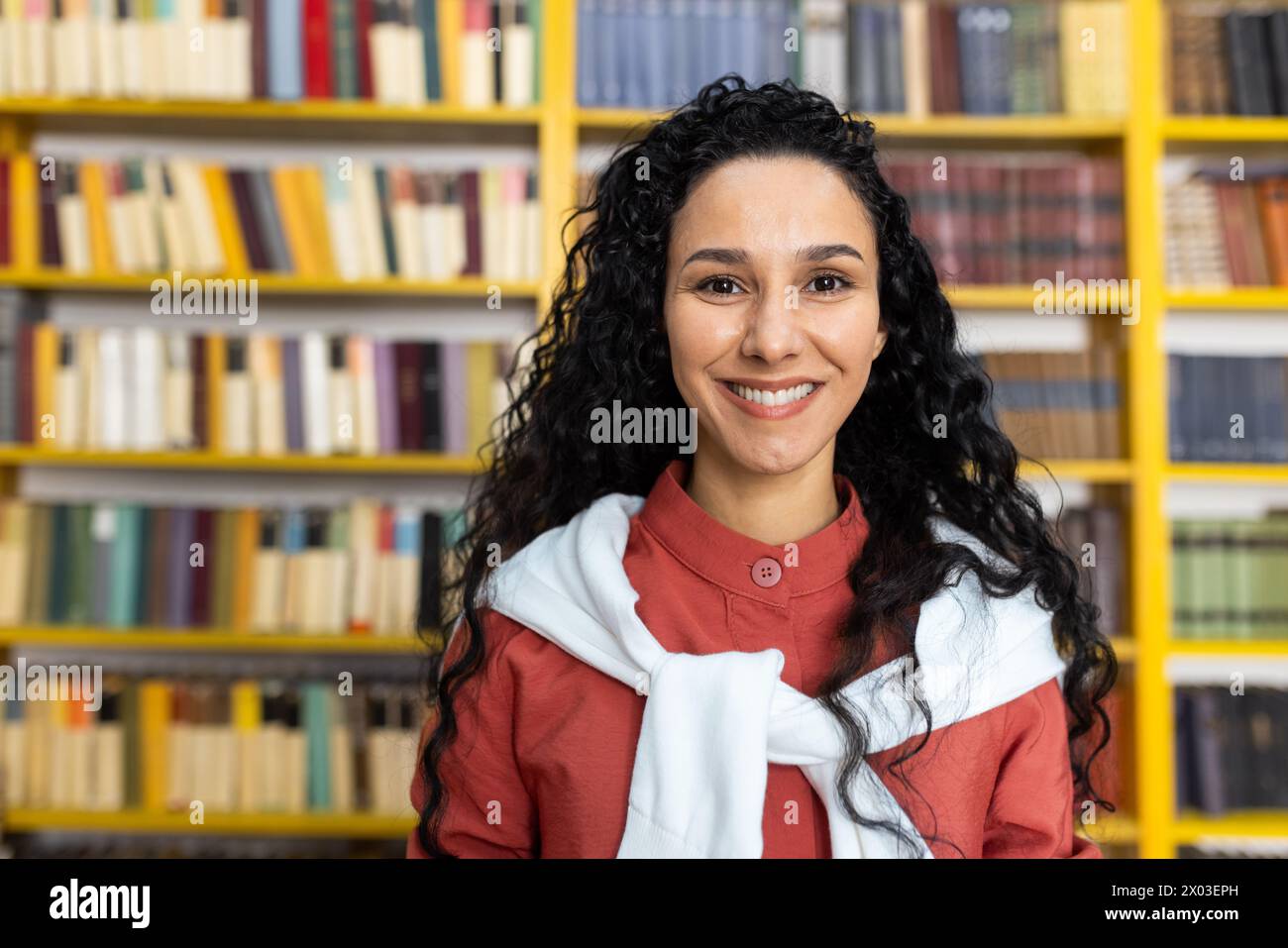 A young Hispanic female student stands in front of a colorful bookshelf ...