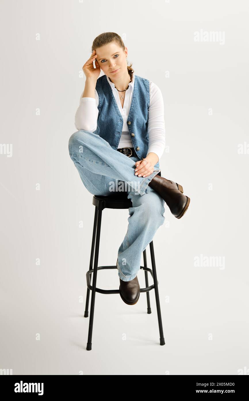 Cute woman in denim outfit sits on tall stool cross-legged on grey ...
