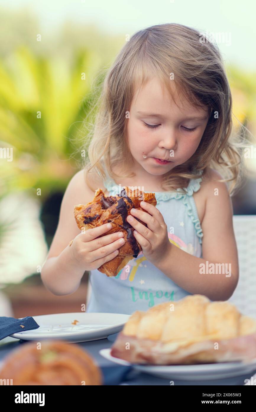 A beautiful little girl eats a croissant with chocolate with appetite ...