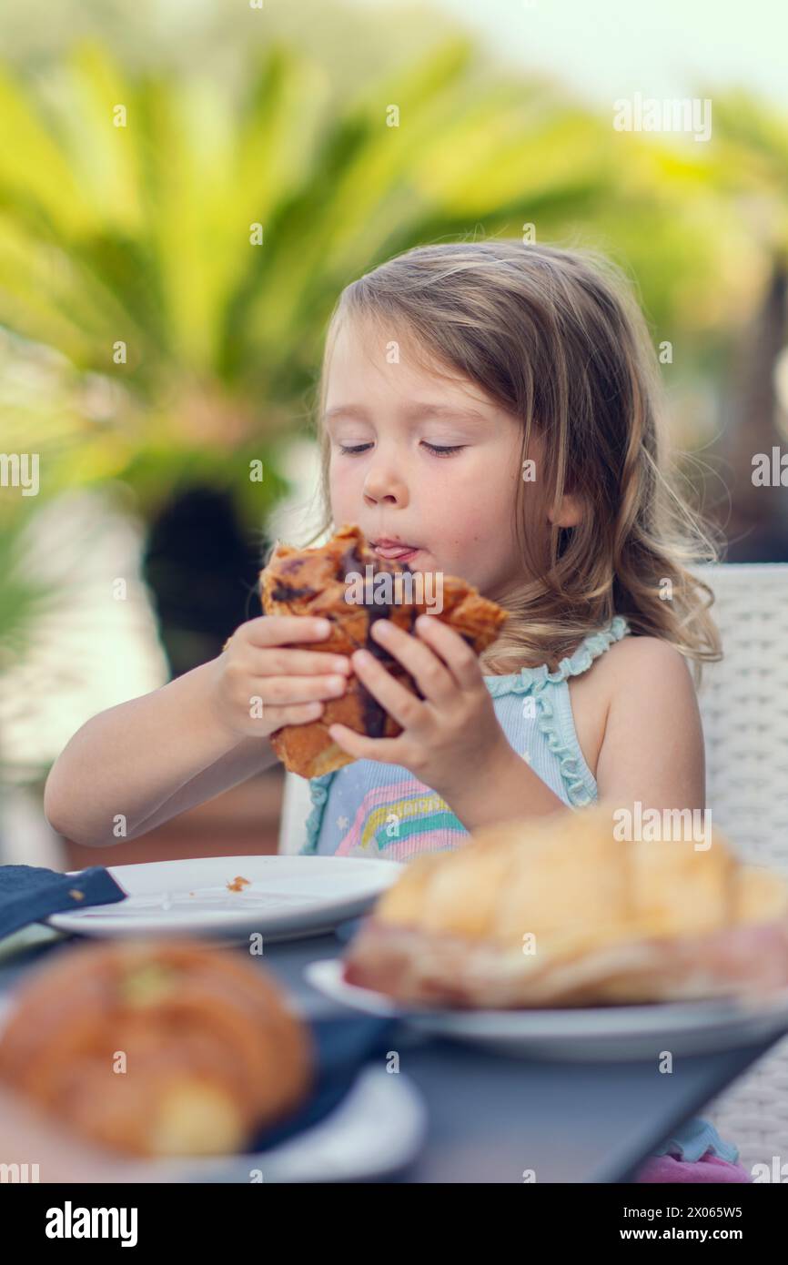 A beautiful little girl eats a croissant with chocolate with appetite ...