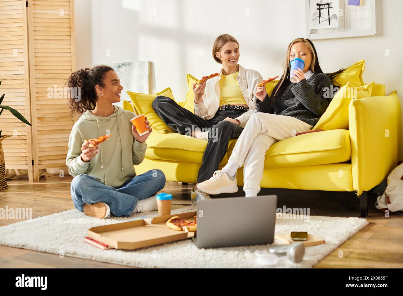 Three diverse teenage girls sit on a yellow couch, bonding over pizza ...