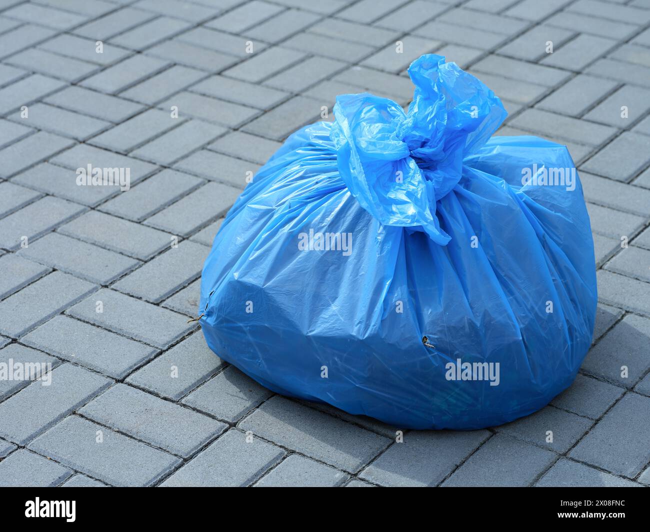 A blue garbage bag full of trash standing on the gray stone paving ...