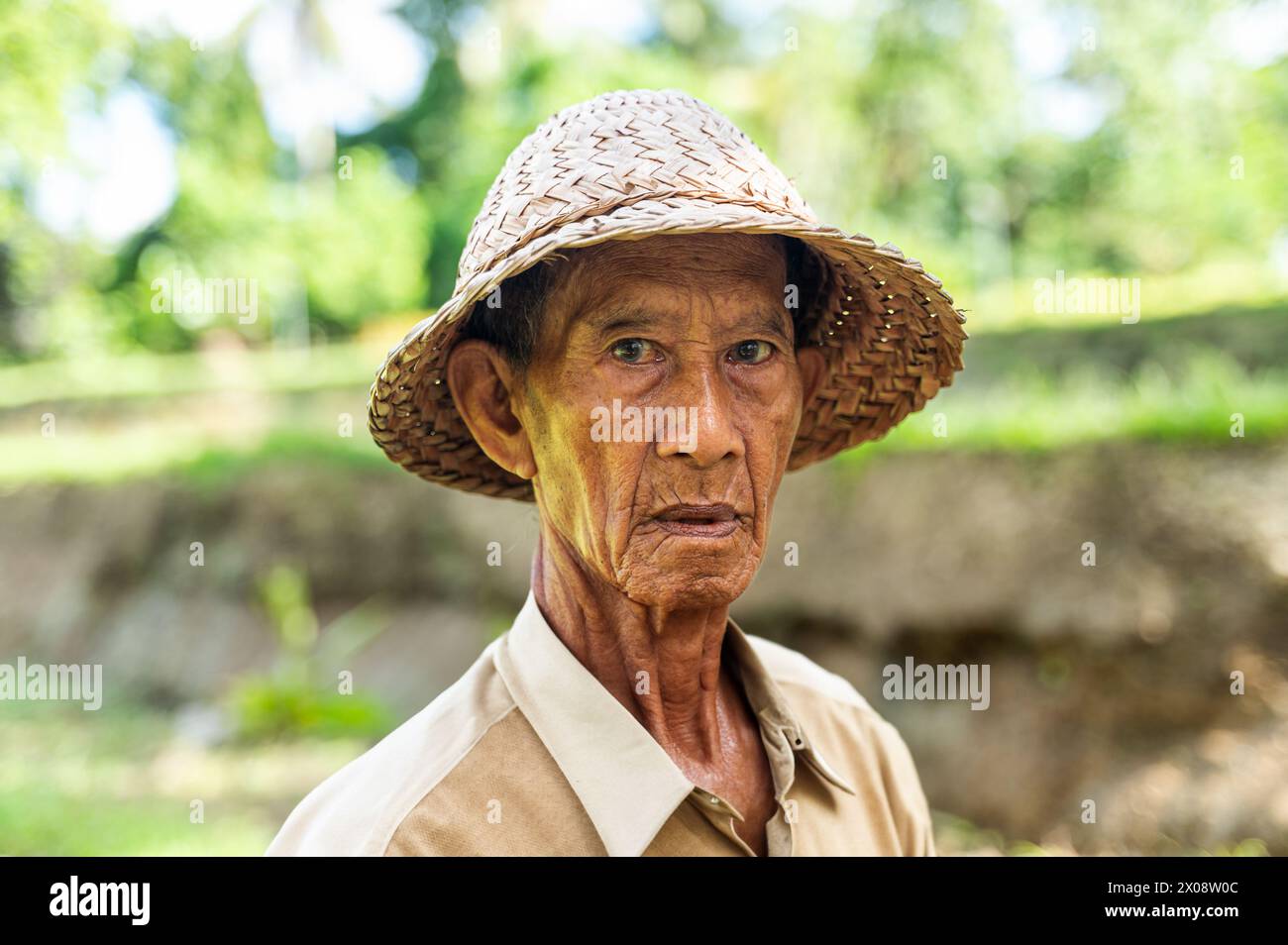 An elderly Balinese man wearing a traditional woven hat stands before a ...