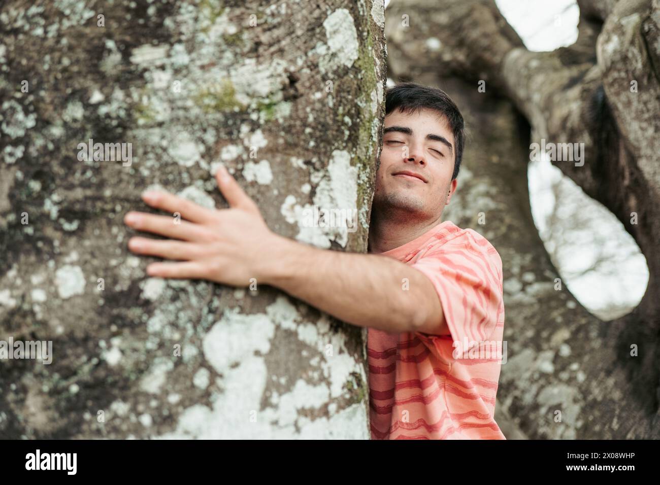 A smiling man joyfully hugs a large, lichen-covered tree, showing a ...