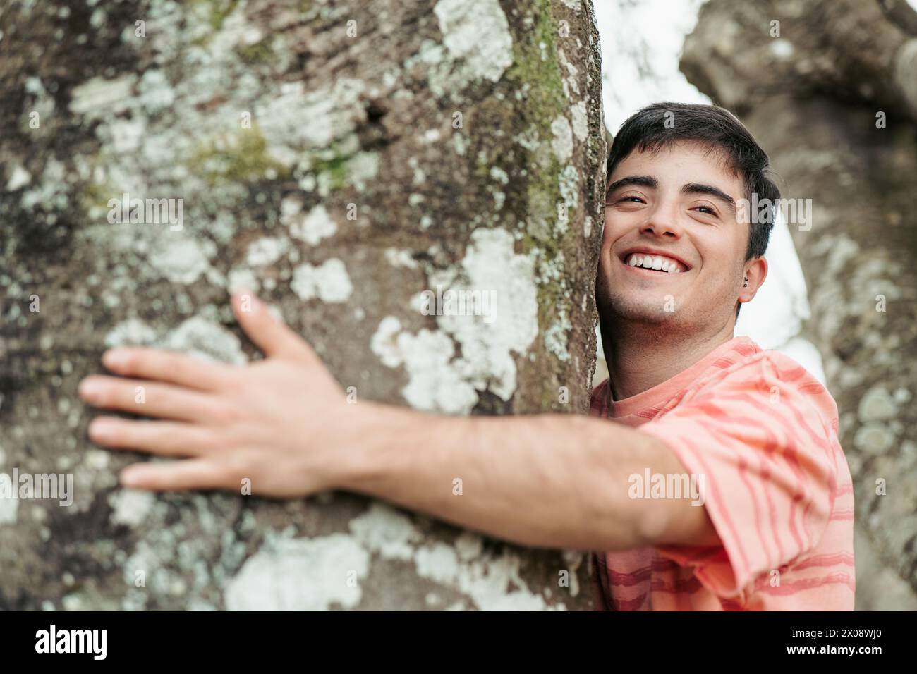 A smiling man joyfully hugs a large, lichen-covered tree, showing a ...