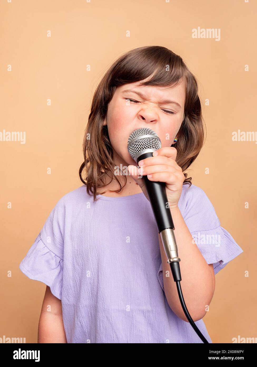 A young girl with a microphone grimaces as she sings passionately against a beige background ...