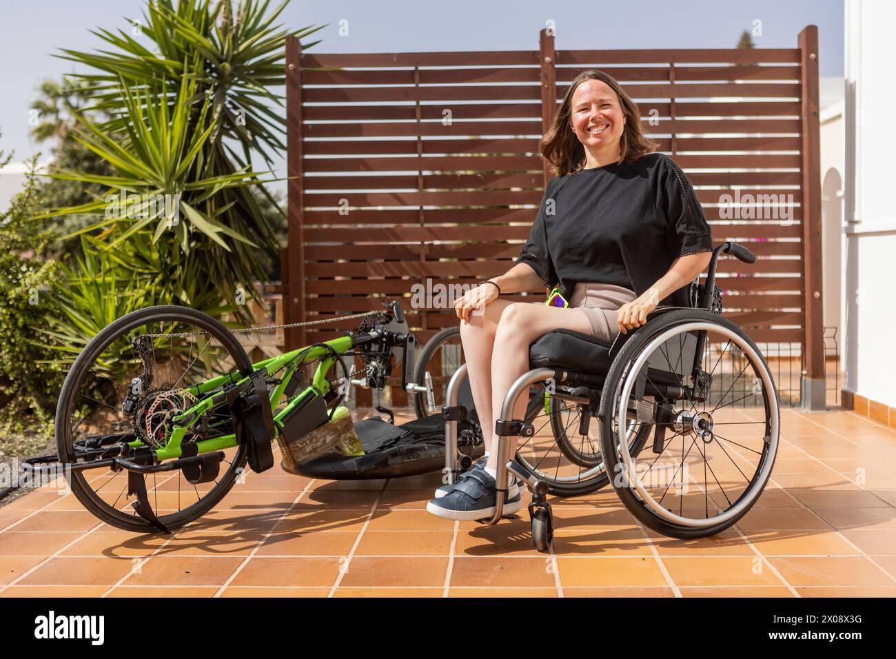 A cheerful woman in sportswear, sitting in a wheelchair next to her racing handbike, exemplifies ...
