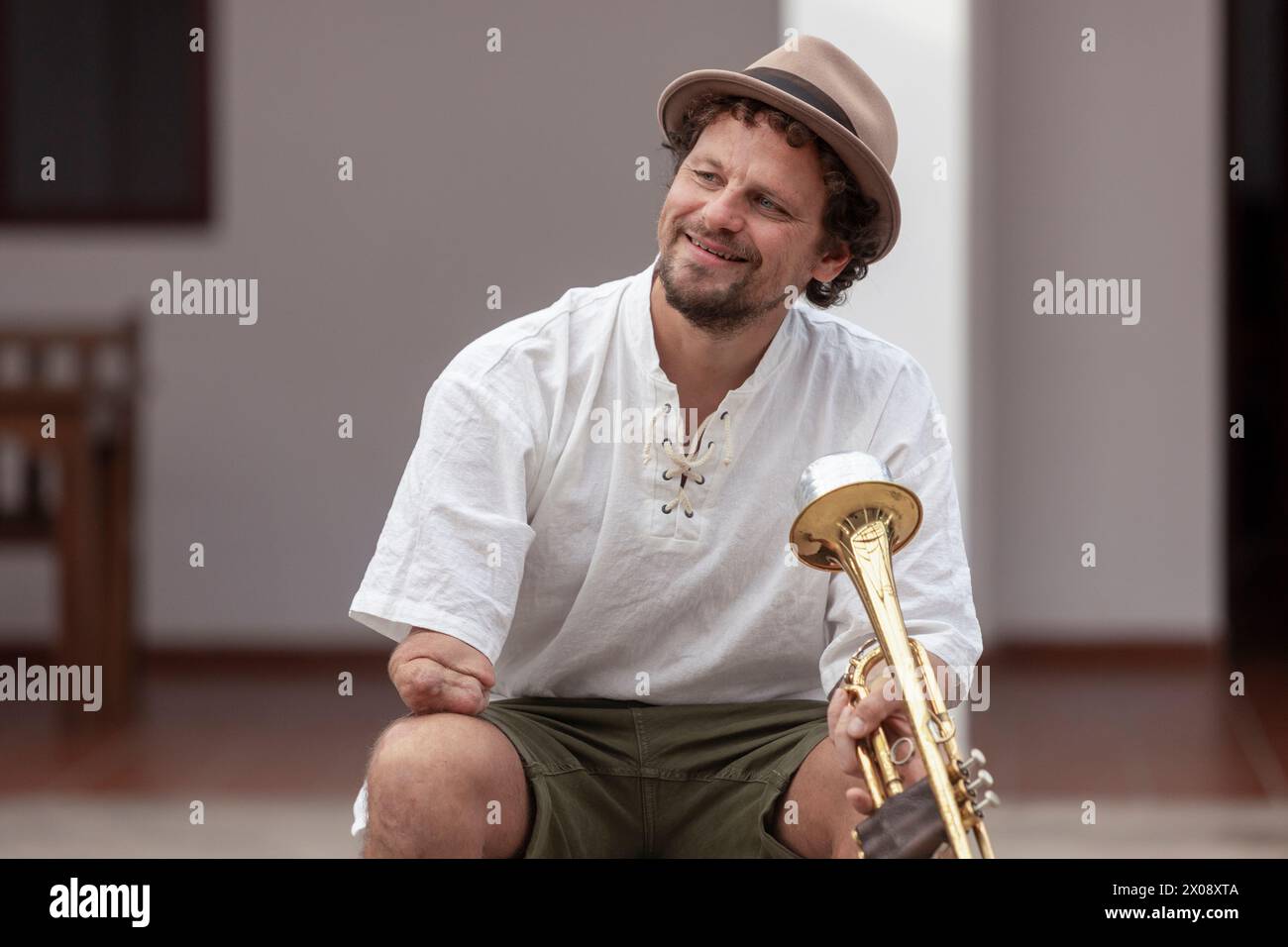 A smiling man with one arm holding a trumpet, sitting in a relaxed pose ...