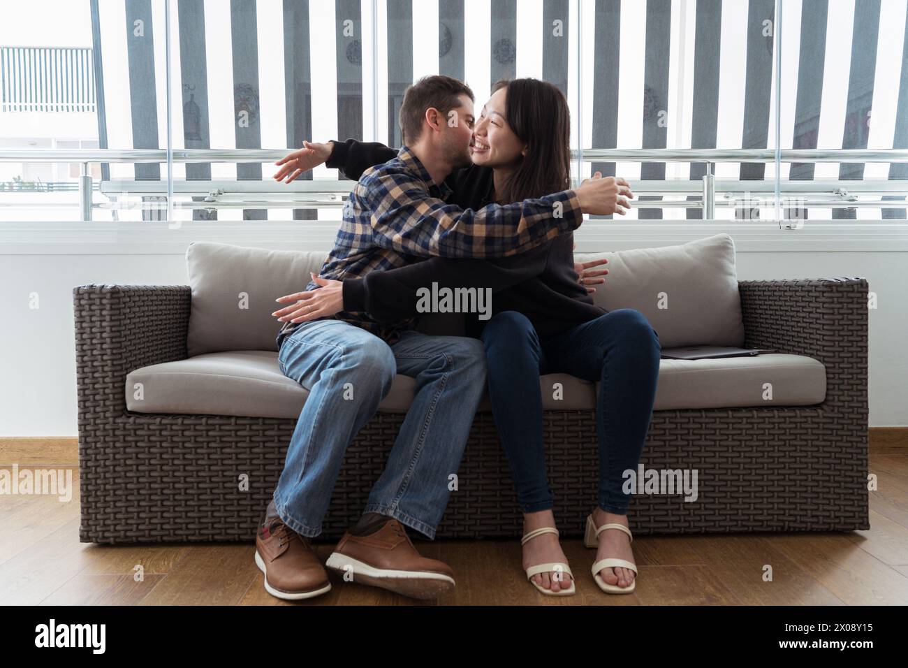 A cheerful Asian woman and a Caucasian man hug on a sofa showcasing ...