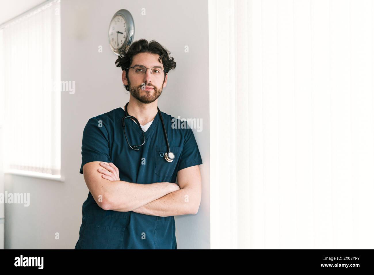 A pensive male healthcare worker with arms crossed stands by a clock ...