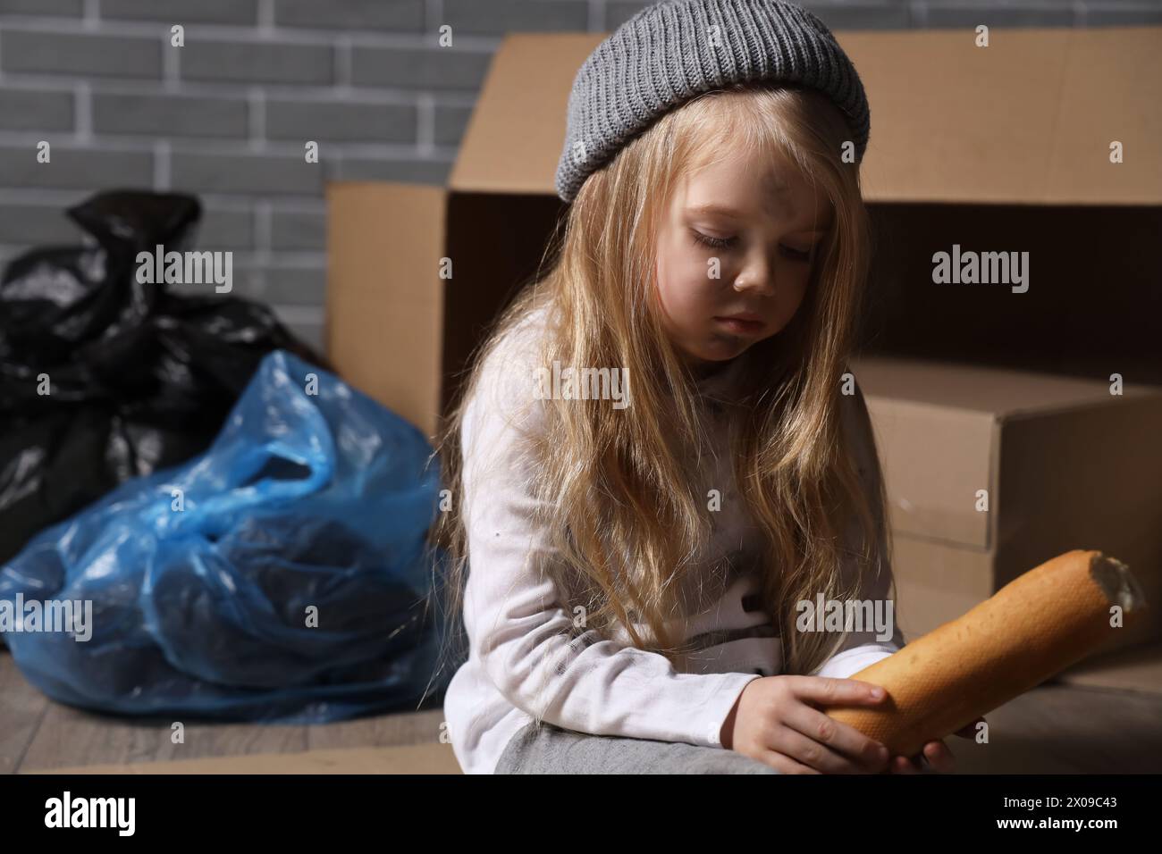 Homeless little girl eating bread near trash against grey brick wall ...