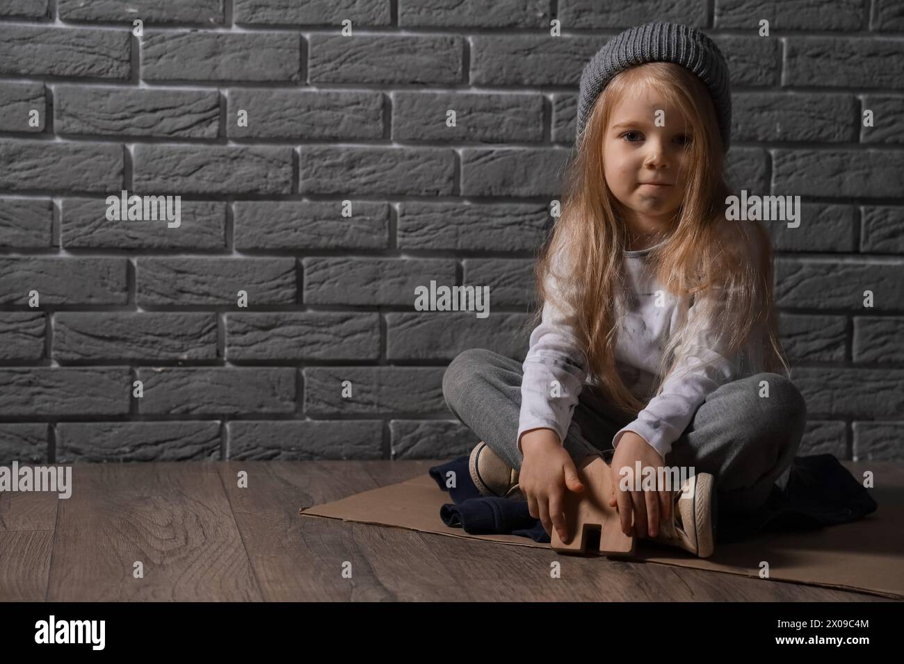 Homeless little girl with figure of house sitting near grey brick wall ...