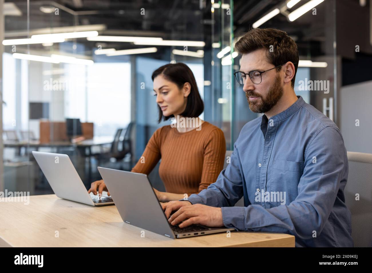 Focused man and woman using laptops at a wooden table in a bright, contemporary workspace ...