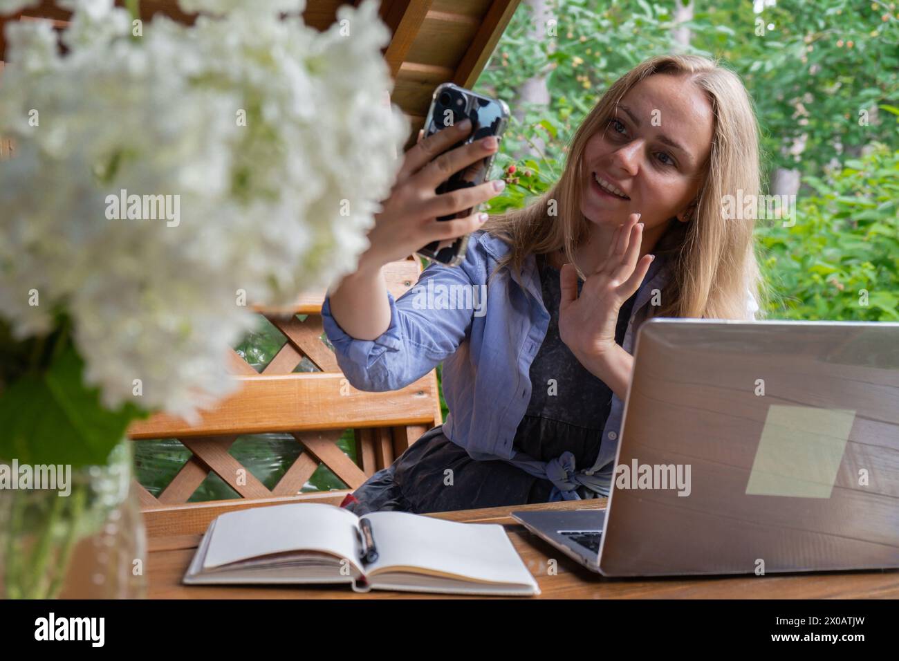 Young happy woman making video call on smartphone in wooden alcove. Relaxed outdoor setting ...