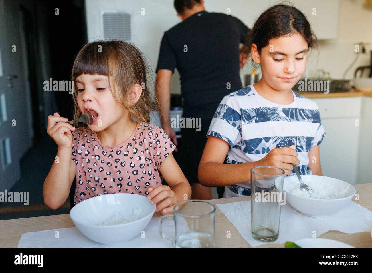 two girls eating lunch at home at white table with father cooking in ...