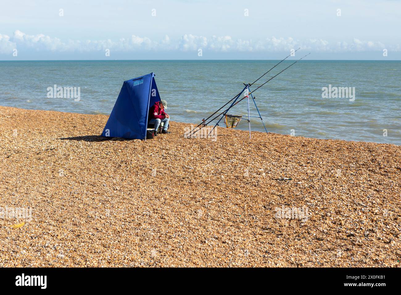 A fisherman in a small windbreak on the shingle beach at Hythe, Kent ...
