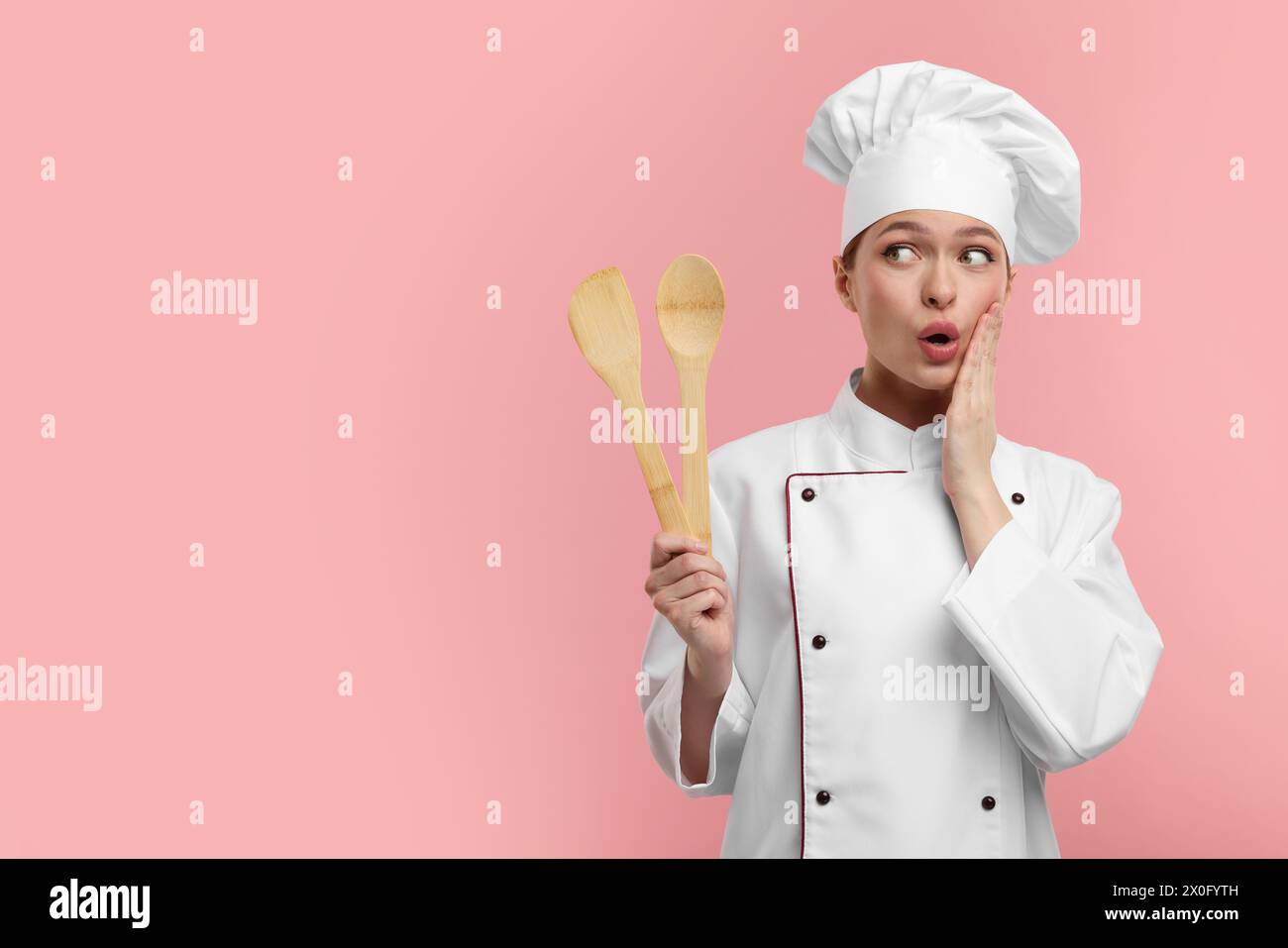 Surprised chef in uniform holding wooden spatula and spoon on pink ...
