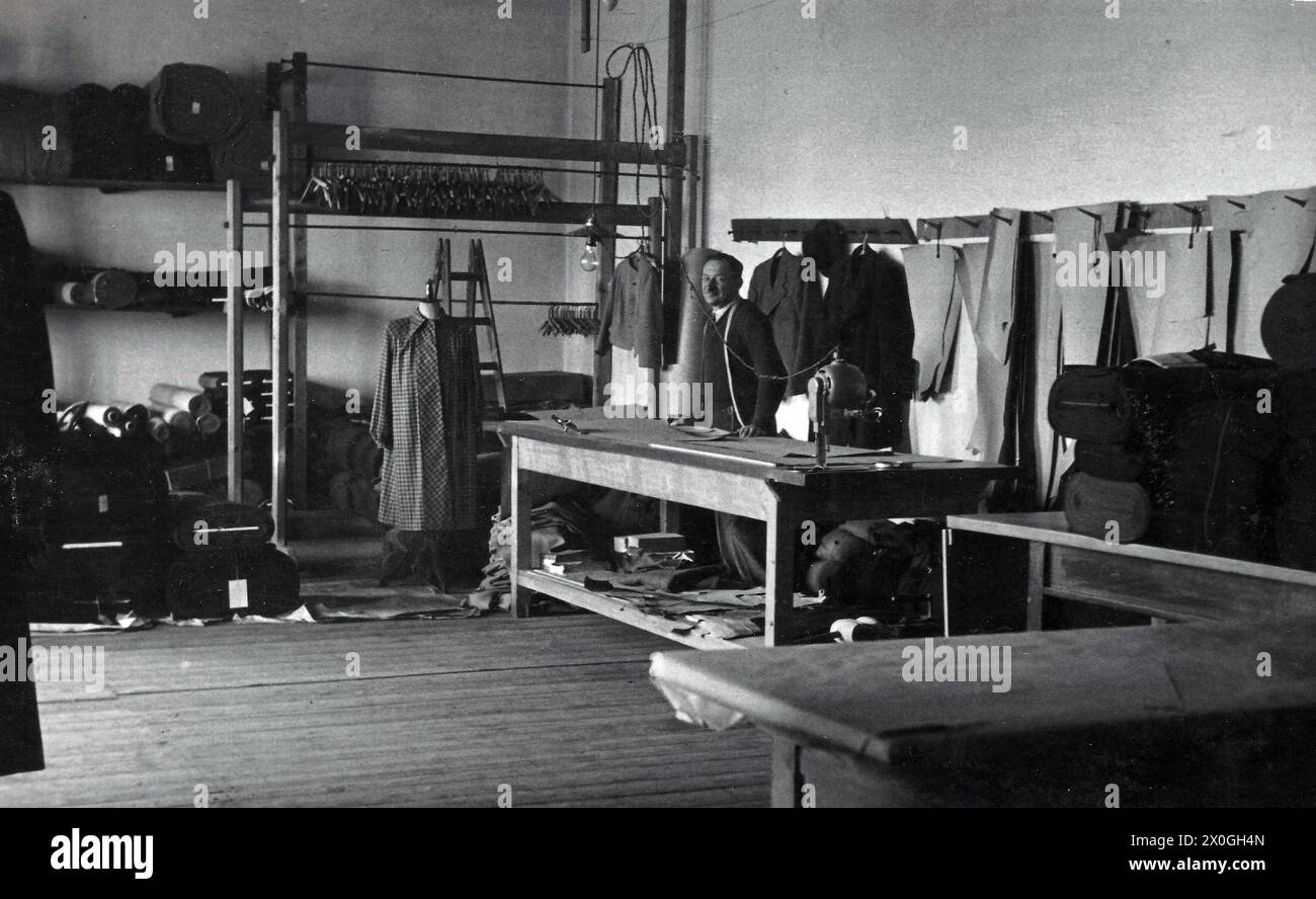 A tailor stands in a large room behind his cutting table surrounded by ...