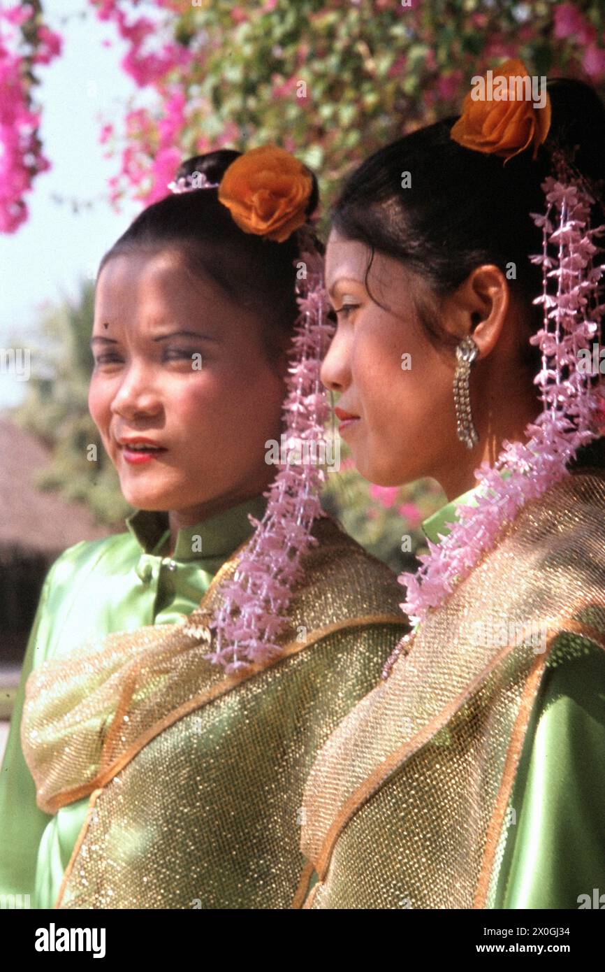 Two Thai Girls In Traditional Clothes And Flowers In Their Hair In A Rose Garden In Bangkok