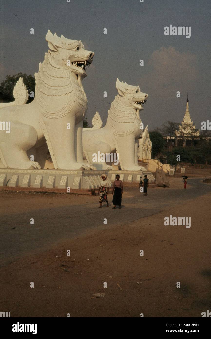 Two women in traditional Myanmar dress pass two large guardian figures ...
