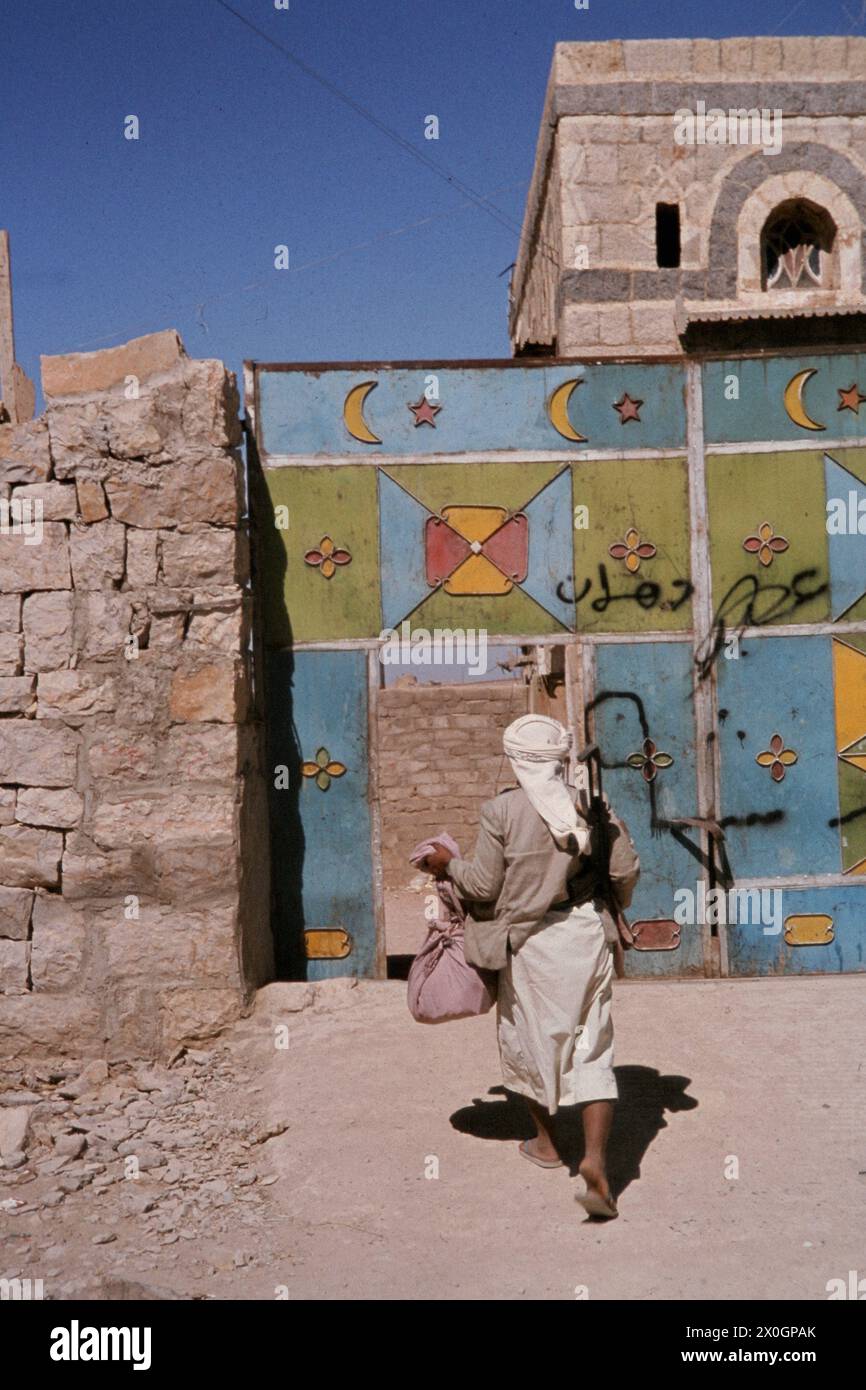 A man with a machine pistol in front of a colourfully painted gate in ...