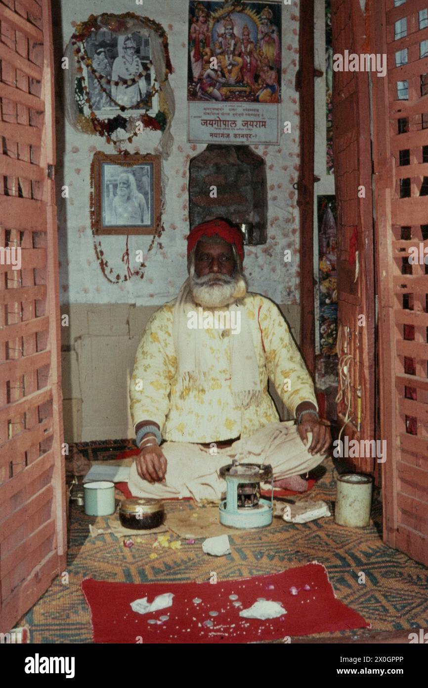 Ein Sadhu mit typischem Vollbart sitzt vor einer Wand mit Bildern im ...