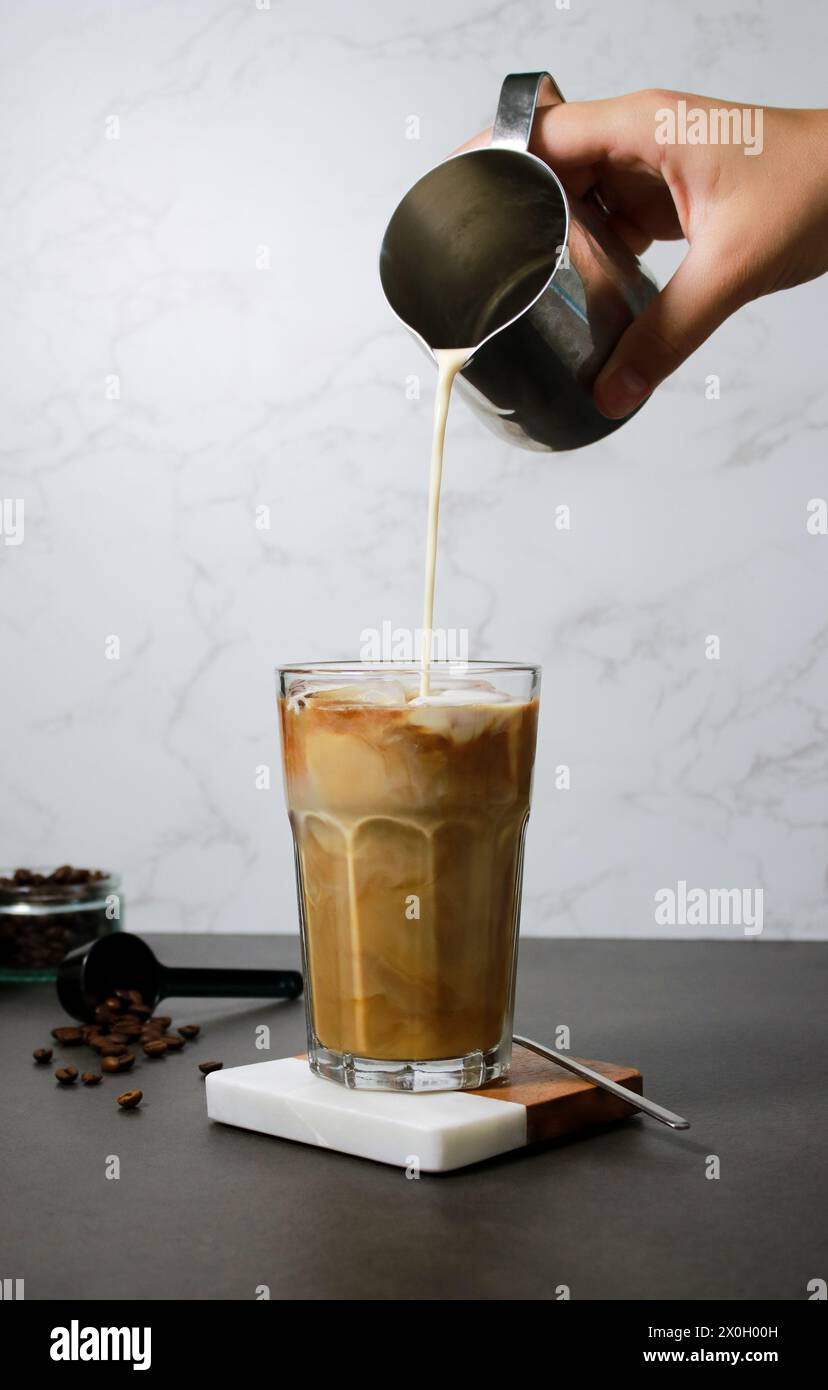 Pouring oat milk into cold ice coffee in a tall glass, grey marble ...