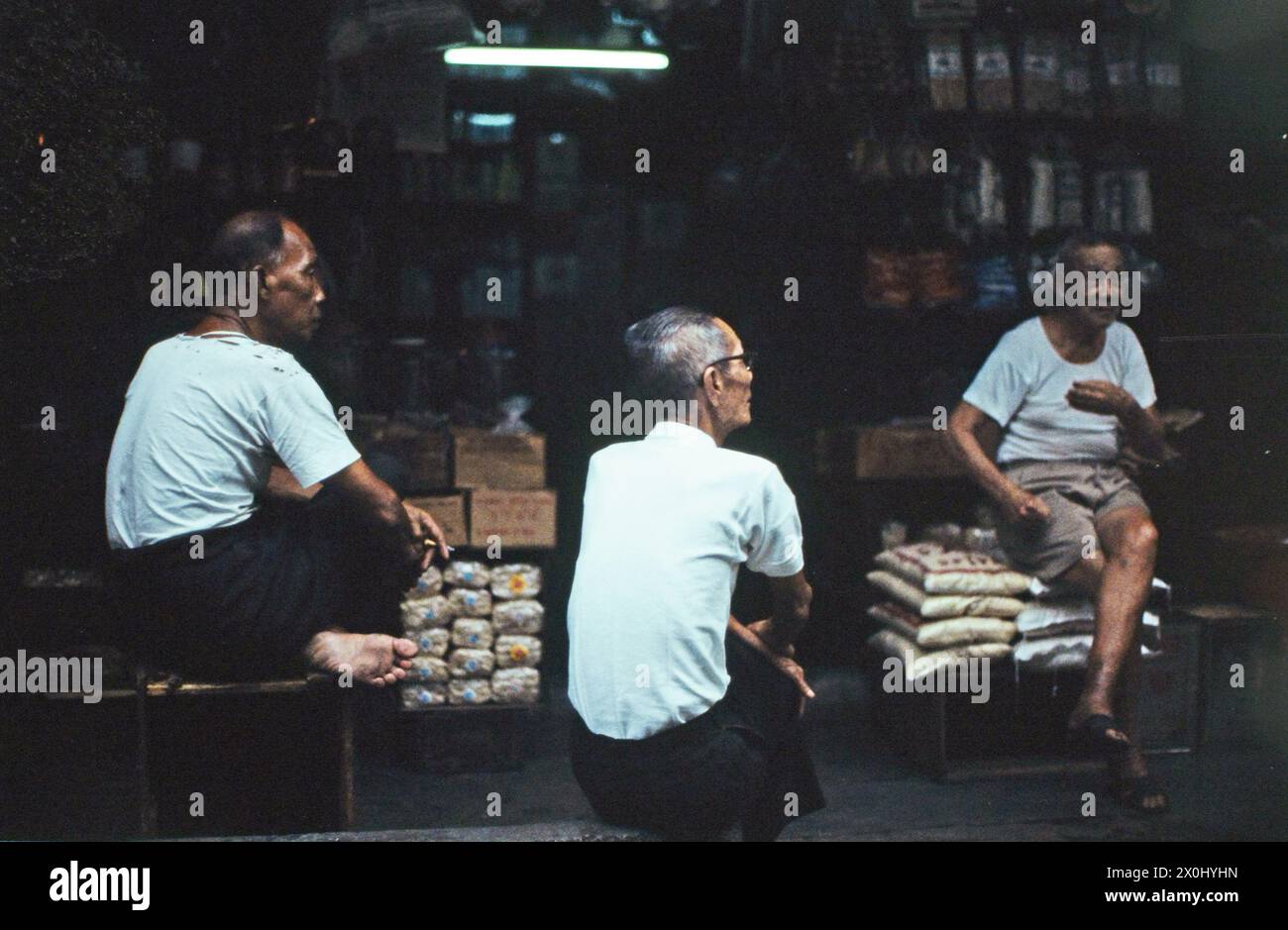 Three older men in turquoise t-shirts are sitting in front of a shop in ...