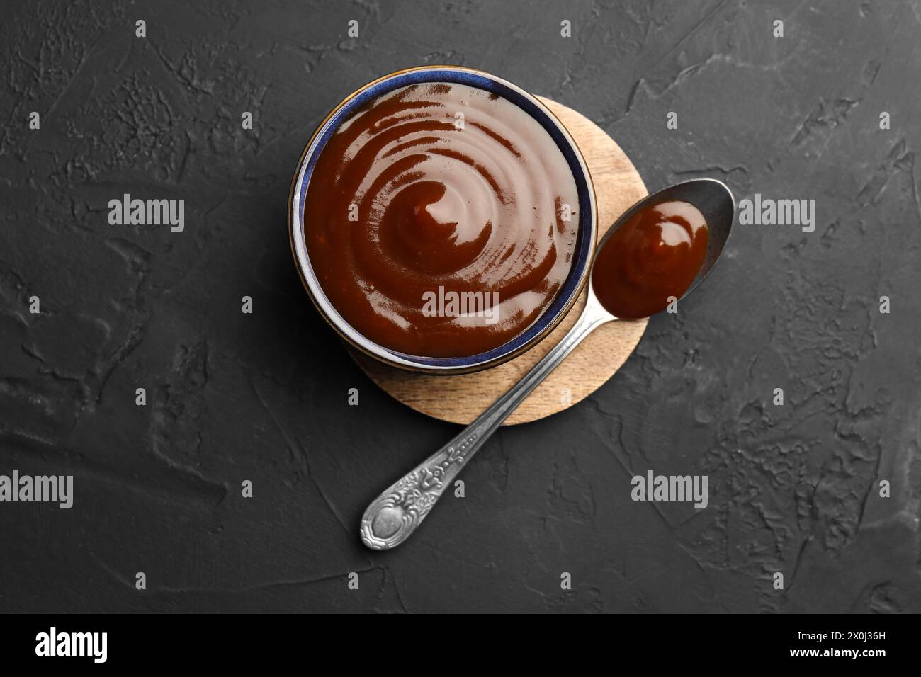 Tasty barbeque sauce in bowl and spoon on dark textured table, top view ...