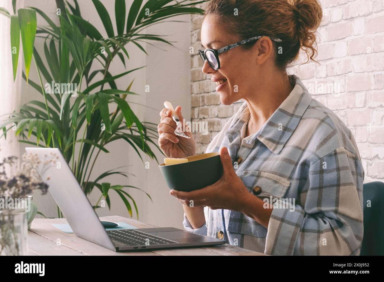 Side view of adult young woman smiling and eating in fron tof a laptop ...