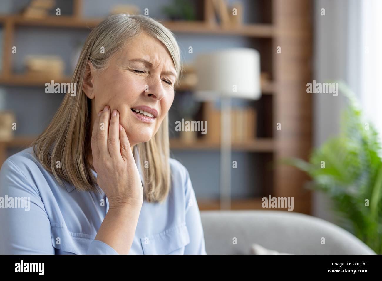 An elderly Caucasian woman sits at home, touching her cheek and ...