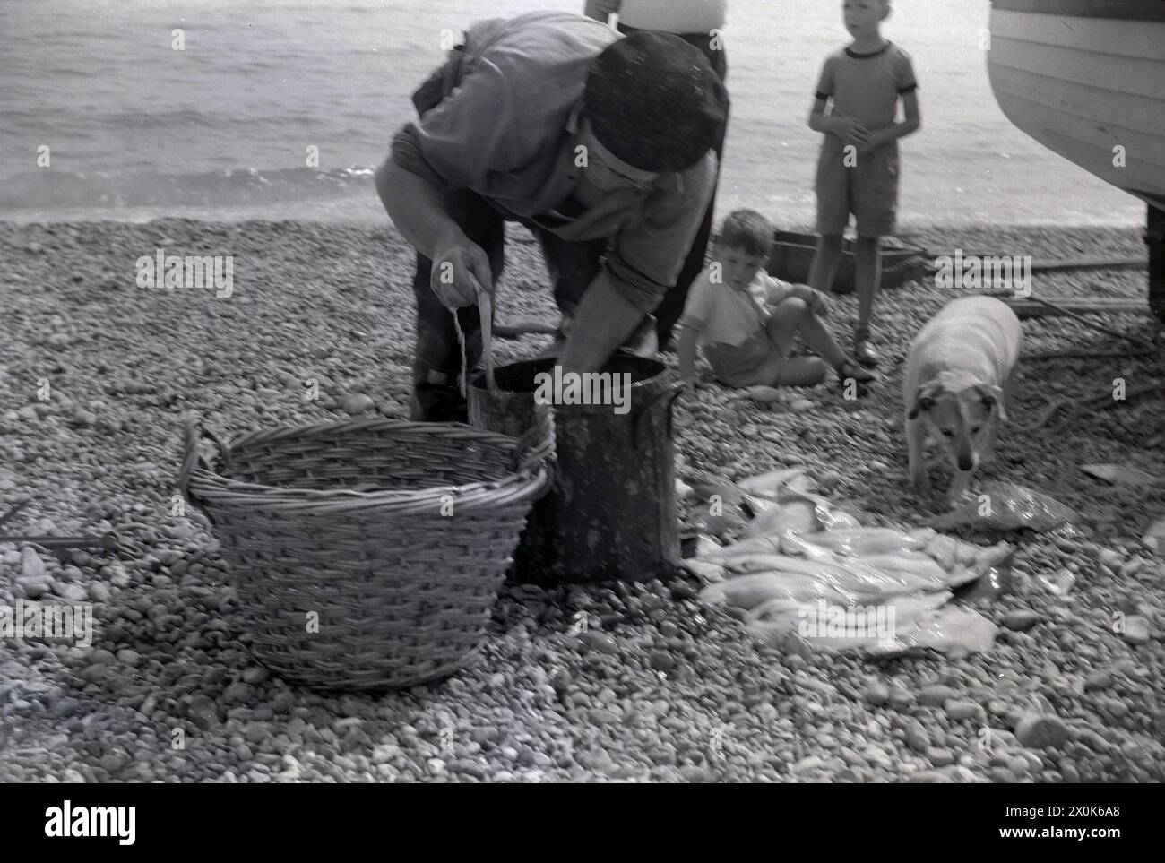 1950s, historical, an elderly fisherman on a beach beside a wicker ...