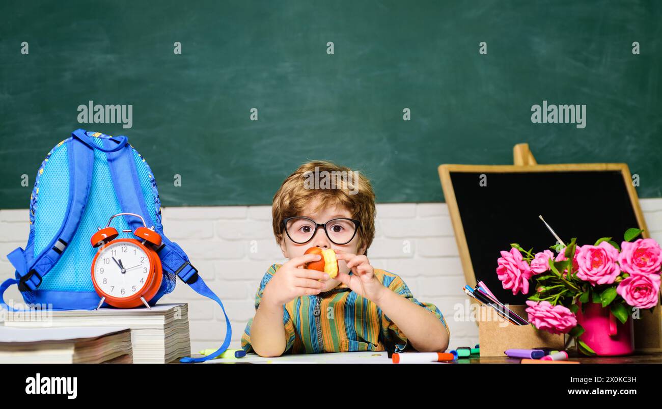 Little schoolboy in glasses at desk in front of chalkboard eating apple ...
