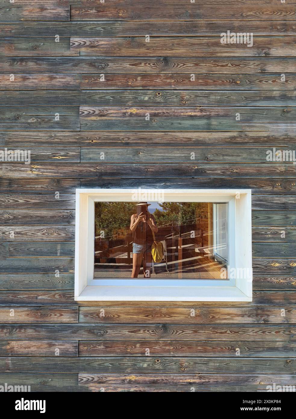 Woman with hat takes a selfie in a small window of a wooden wall as a ...